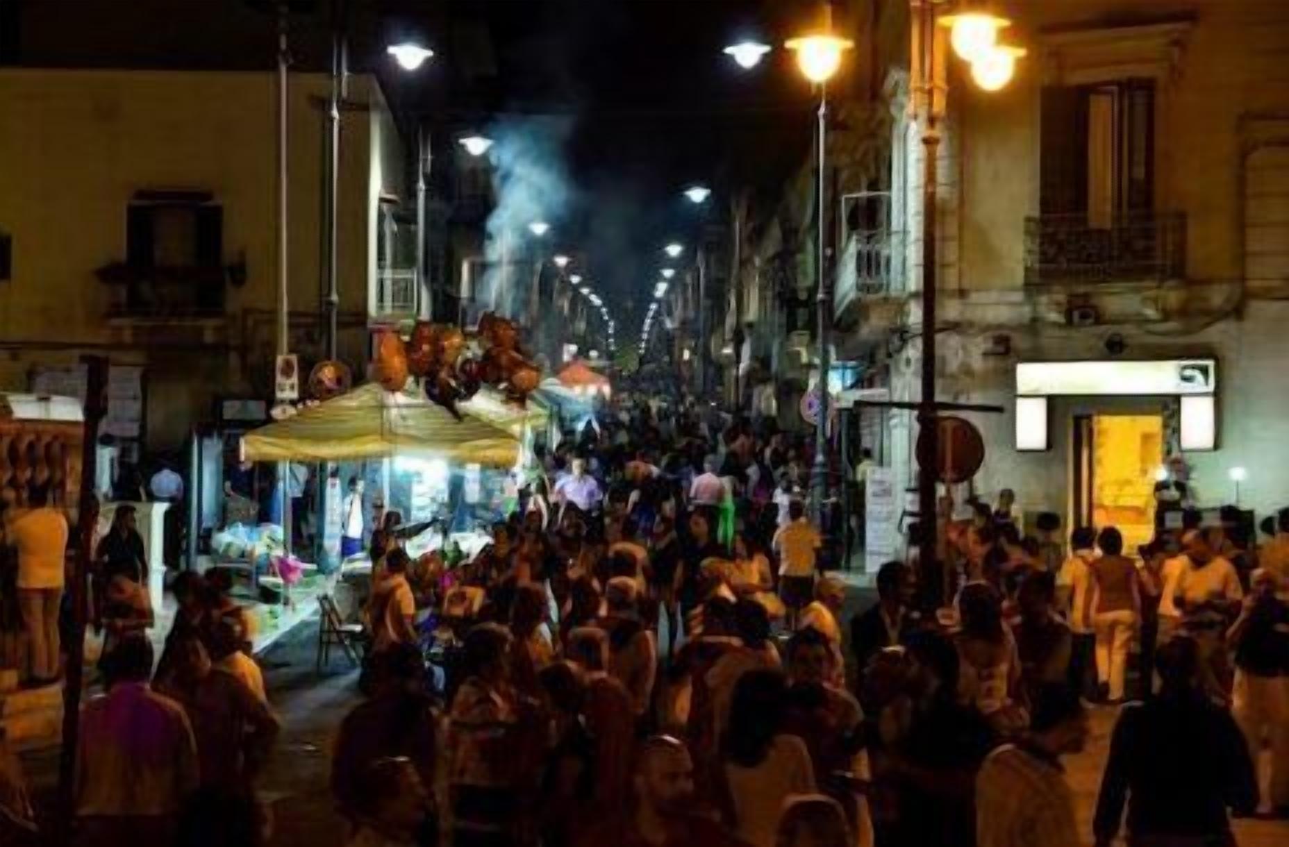 Cena de rua movimentada à noite, com pessoas reunidas em torno de uma área central, possivelmente um mercado ou evento.