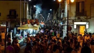 Cena de rua movimentada à noite, com pessoas reunidas em torno de uma área central, possivelmente um mercado ou evento.