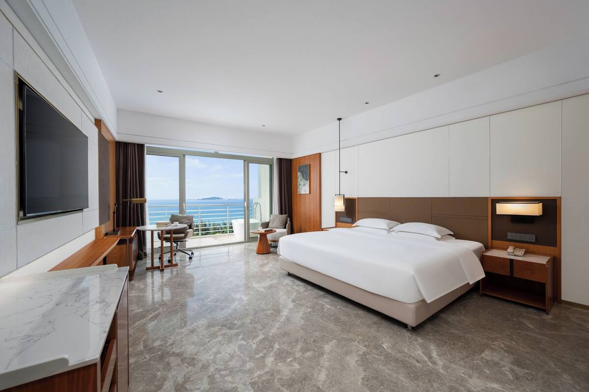Quarto de hotel moderno com uma cama grande, uma escrivaninha com cadeira, televisão e vista para o oceano.