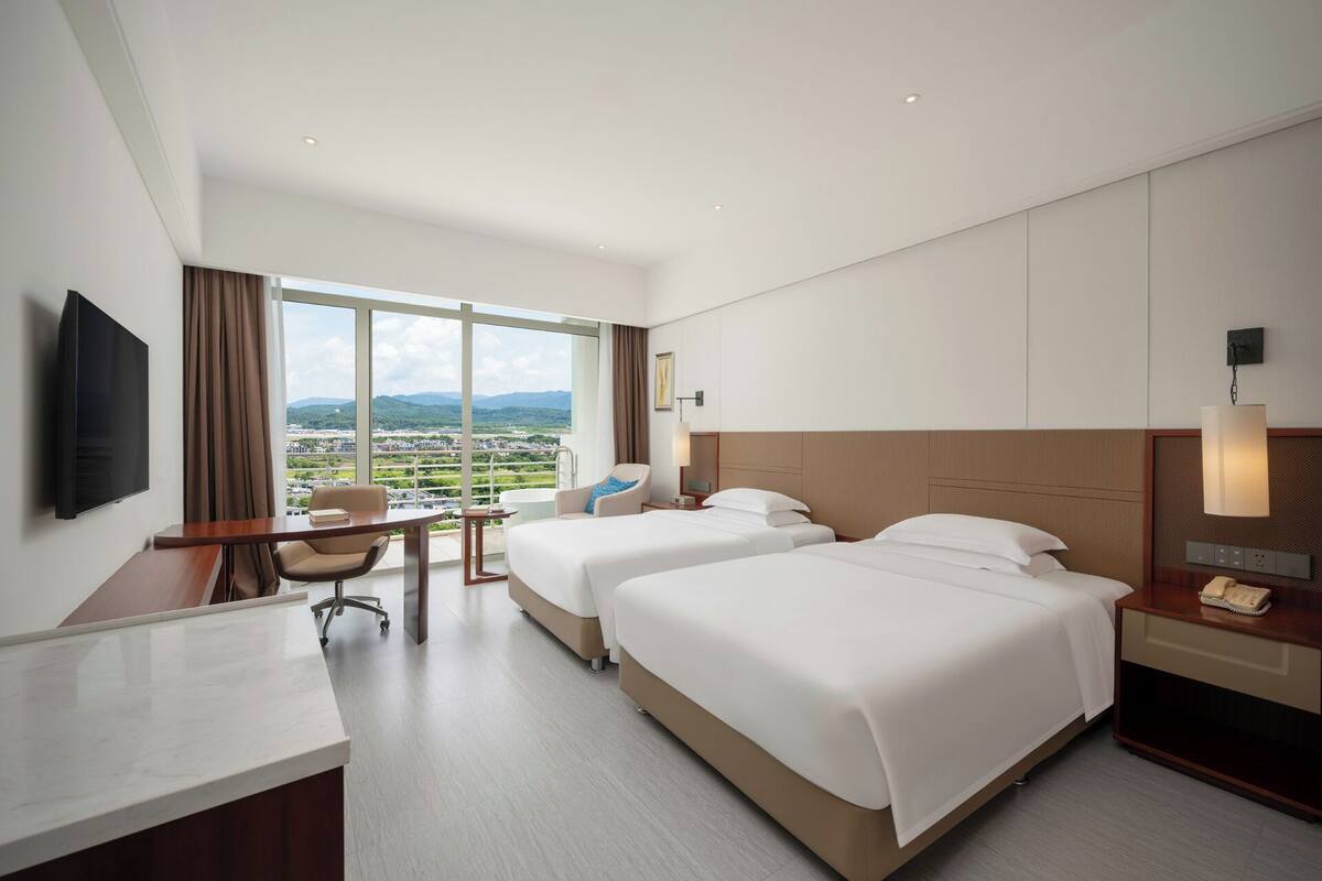 Quarto de hotel com duas camas, uma escrivaninha, uma cadeira, televisão e vista para a cidade.