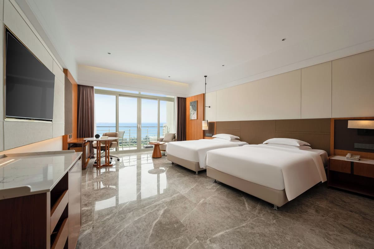Quarto de hotel com duas camas, uma escrivaninha, televisão e varanda com vista.