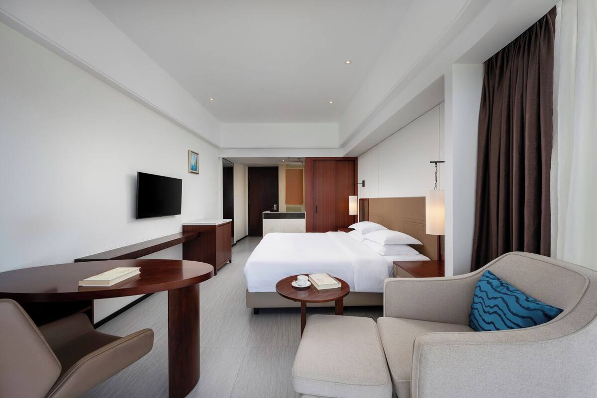 Quarto de hotel com cama, escrivaninha, cadeira, mesa pequena e televisão.