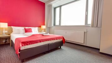 Quarto de hotel com cama, duas luminárias de cabeceira, janela com cortinas e radiador.