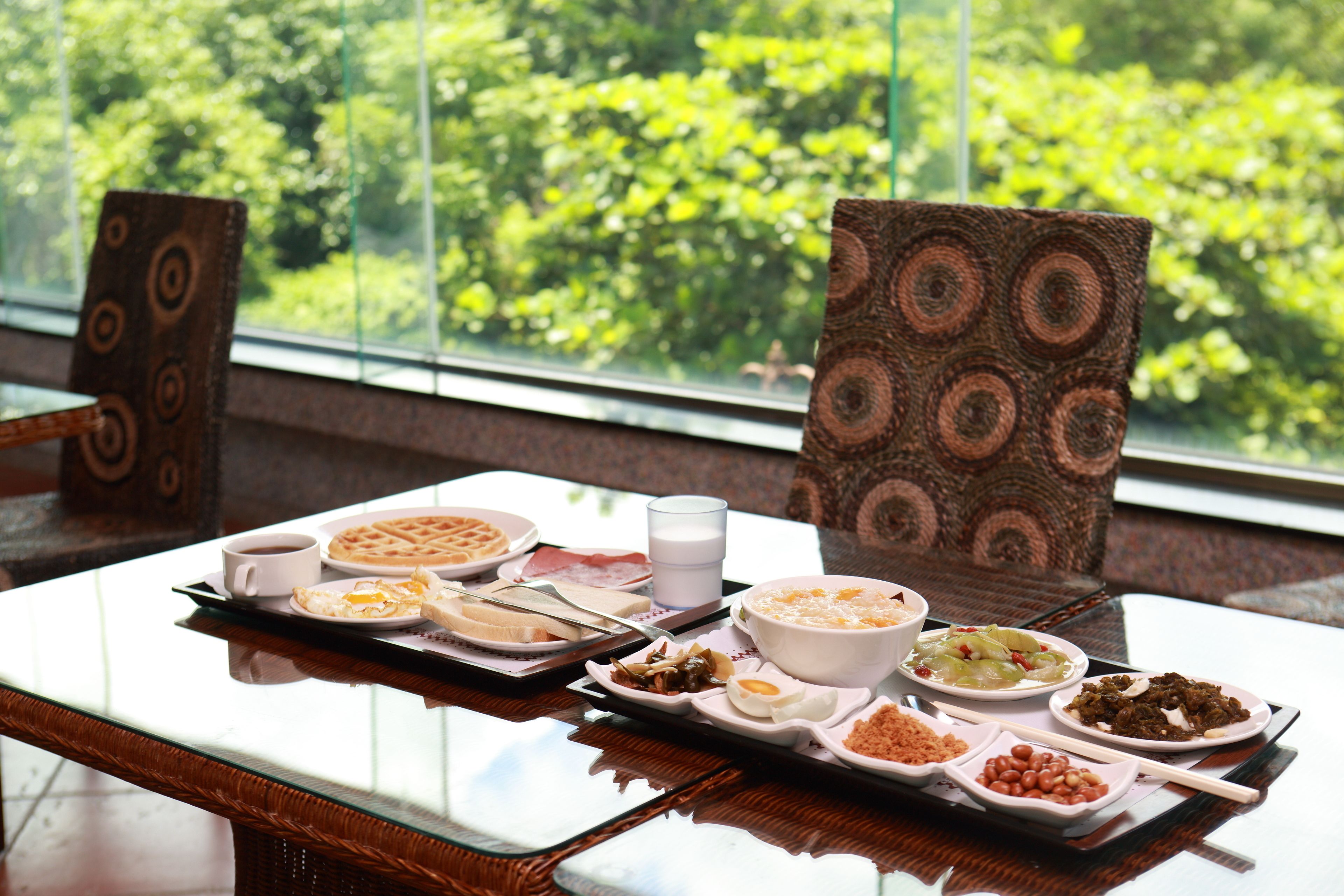 Mesa de café da manhã com diversos pratos e um copo de leite, em frente a uma janela com vista para o verde.