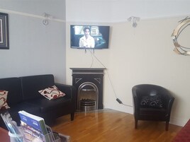 Uma sala de estar com um sofá de couro preto, uma poltrona preta, uma lareira e uma televisão fixada na parede.