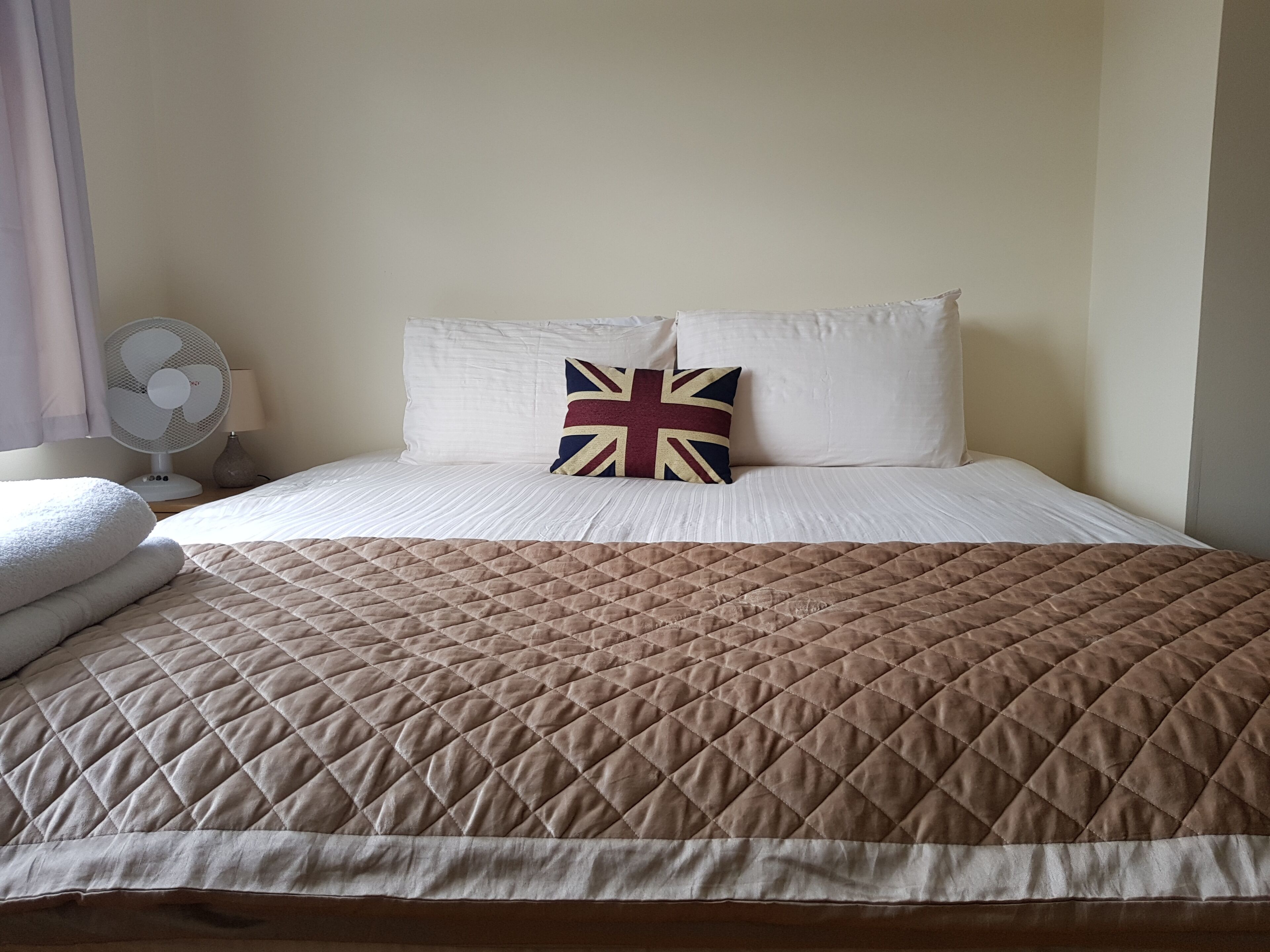 Cama bem arrumada com cobertura marrom acolchoada, almofadas brancas e uma almofada com a bandeira do Reino Unido.