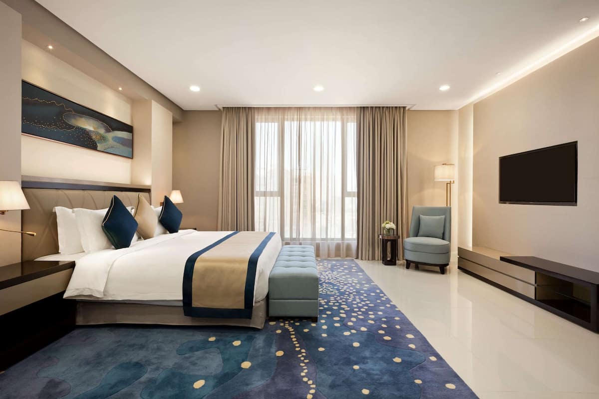 Quarto de hotel moderno com uma cama grande, área de estar, televisão e vista da cidade através de amplas janelas.