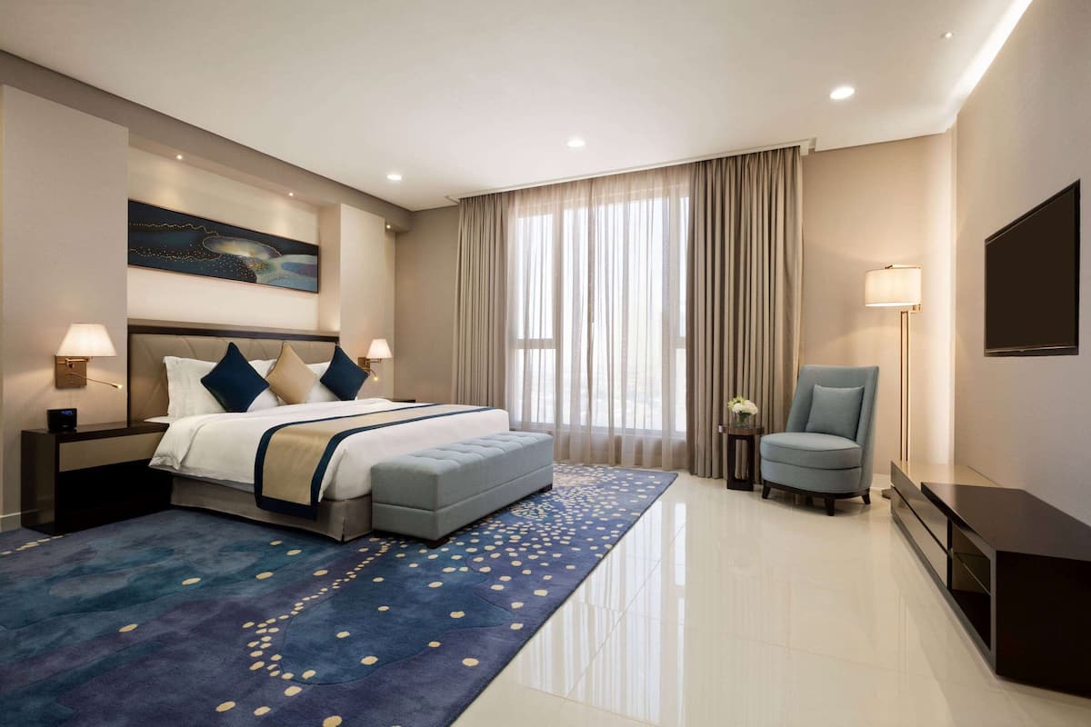 Quarto de hotel moderno com uma cama grande, área de estar, televisão de tela plana e uma janela ampla com cortinas.