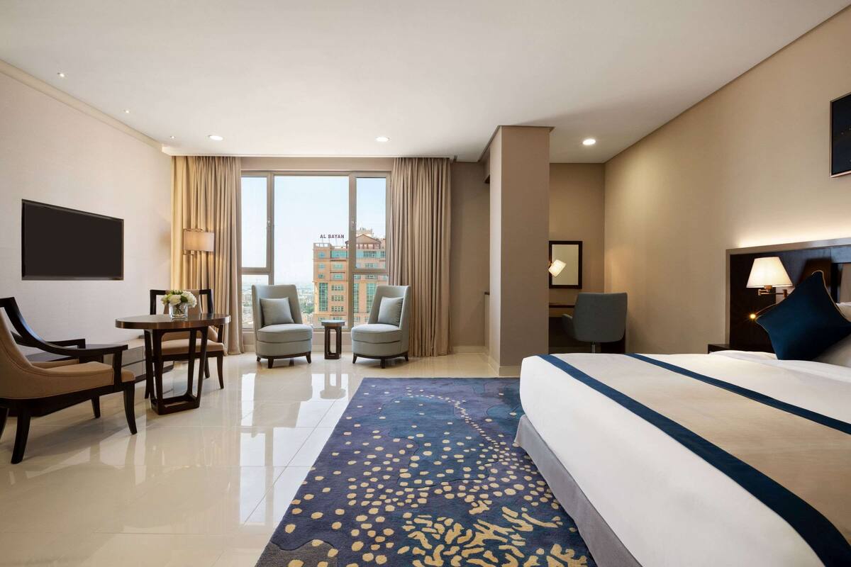 Quarto de hotel com uma cama grande, duas poltronas, uma mesa pequena com um vaso, uma luminária e vista para a paisagem urbana.