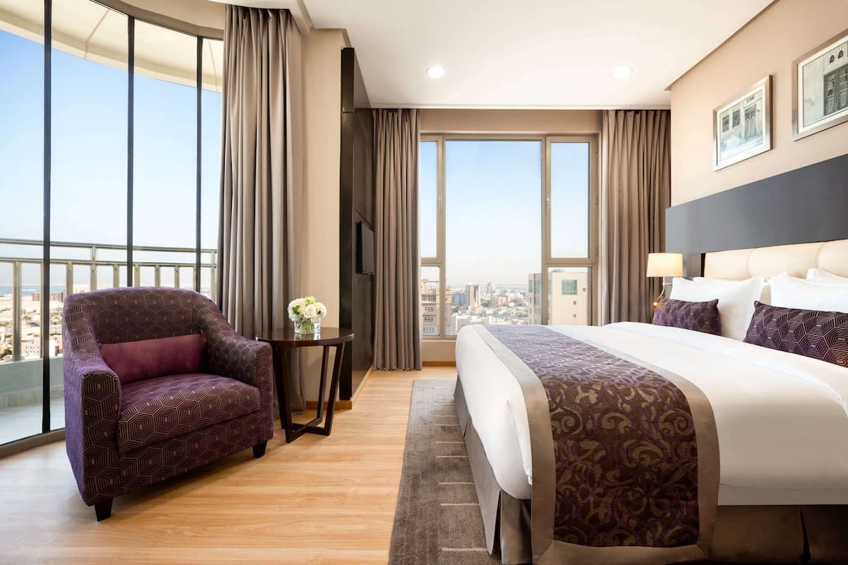 Quarto de hotel com uma cama grande, um sofá, uma pequena mesa com um vaso e vista da cidade através de amplas janelas.