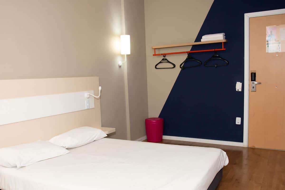 Quarto de hotel com cama, luminária na parede, cabideiro, porta e lixeira.