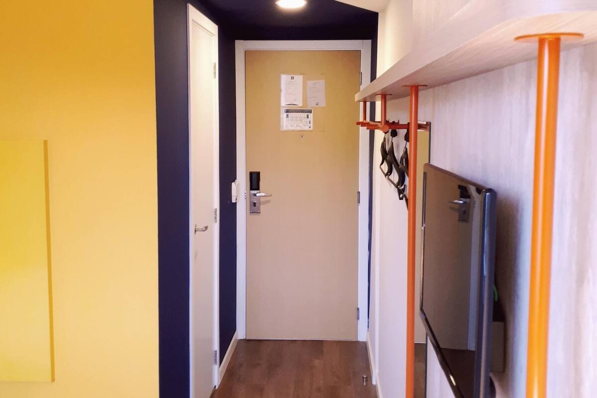 Um corredor com piso de madeira, parede amarela, porta branca e uma televisão fixada na parede à direita.
