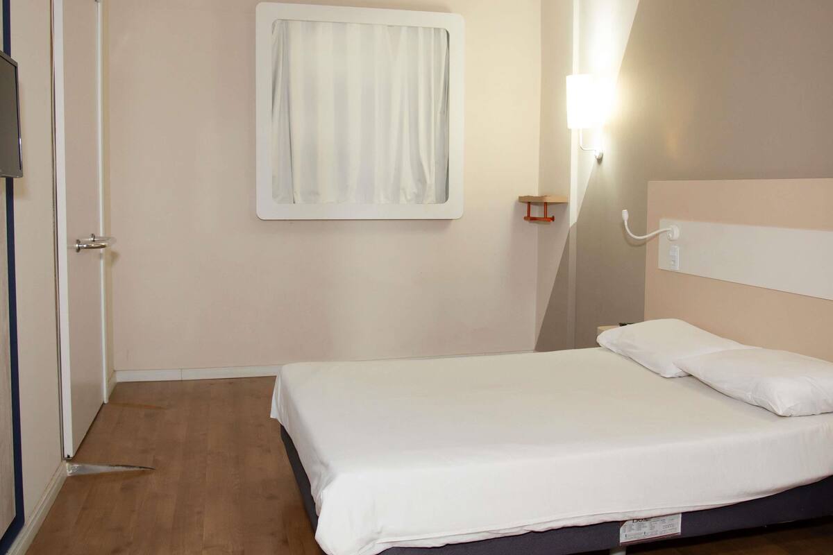 Quarto de hotel com cama, janela com cortinas, luminária na parede e porta.