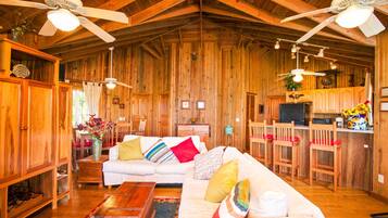 Uma sala de estar espaçosa com paredes e teto de madeira, mobiliada com dois sofás brancos, uma mesa de centro e um canto de jantar com cadeiras de madeira e uma pequena cozinha.