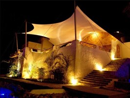 Um edifício moderno, com vários níveis, destacando-se por uma cobertura branca imponente, uma escadaria que leva a um andar superior e uma área de piscina na base.