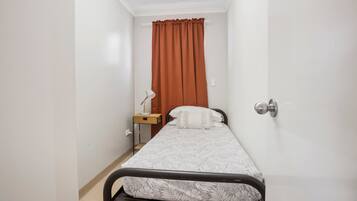 Um quarto pequeno e bem organizado, com uma cama de solteiro, mesa de cabeceira com abajur e janela com cortinas.