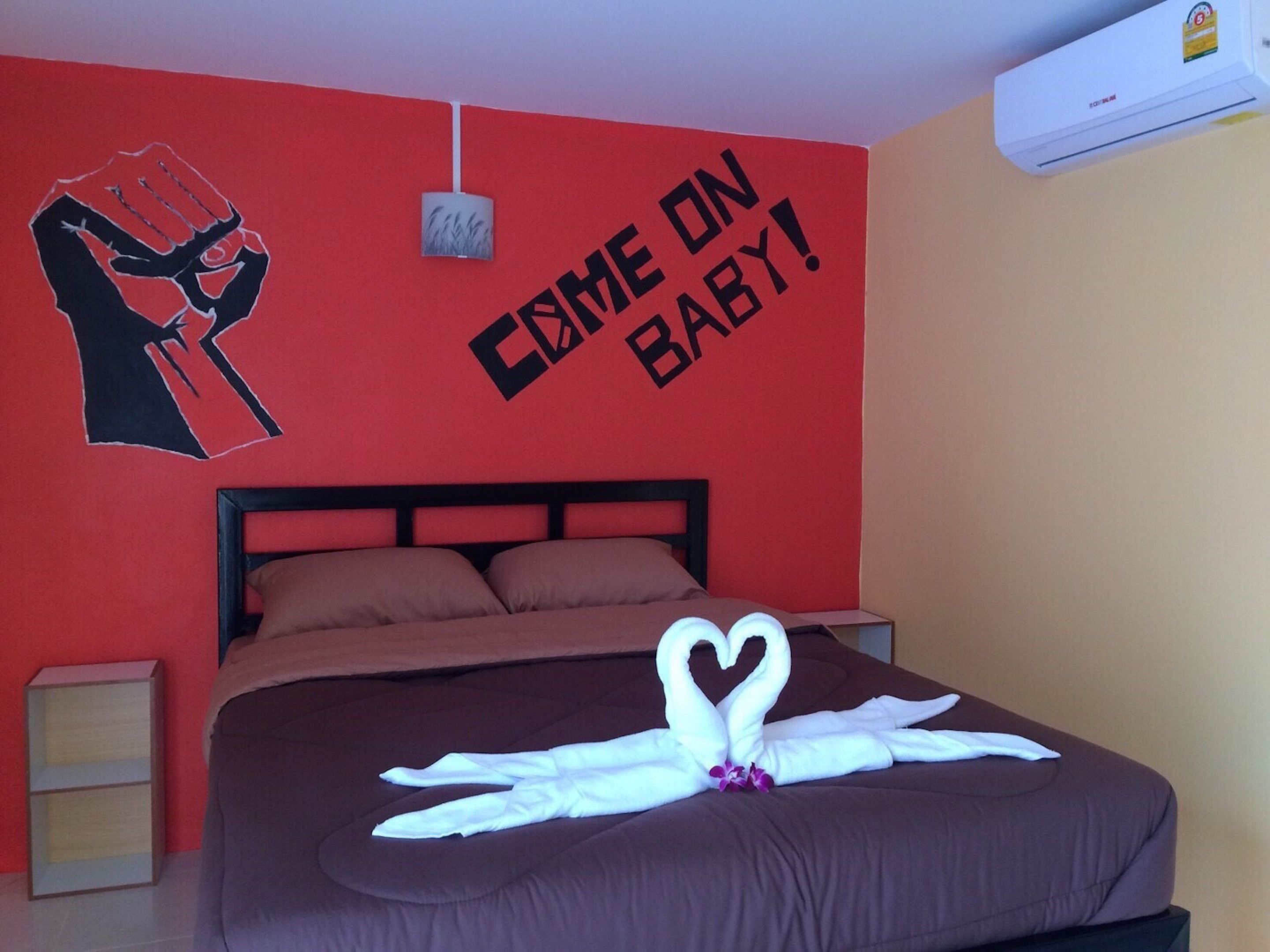 Quarto de hotel com parede vermelha, cama com roupa de cama marrom e decoração de parede com um punho cerrado e o texto "COME ON BABY!".