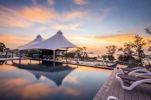 Uma área de piscina tranquila com duas toldas brancas, um deck de madeira e vista para o pôr do sol sobre a água.
