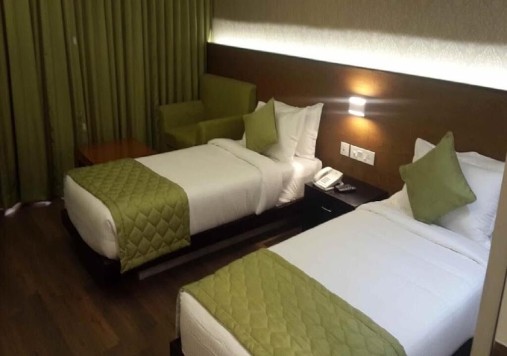 Quarto de hotel com duas camas, cada uma com colcha verde acolchoada e almofadas brancas. Tem uma mesinha de cabeceira com um telefone e uma lâmpada.