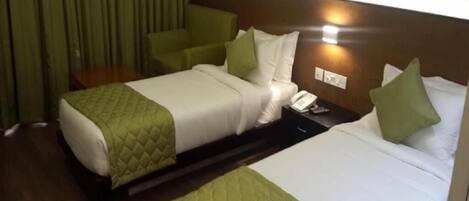 Quarto de hotel com duas camas, cada uma com colcha verde acolchoada e almofadas brancas. Tem uma mesinha de cabeceira com um telefone e uma lâmpada.