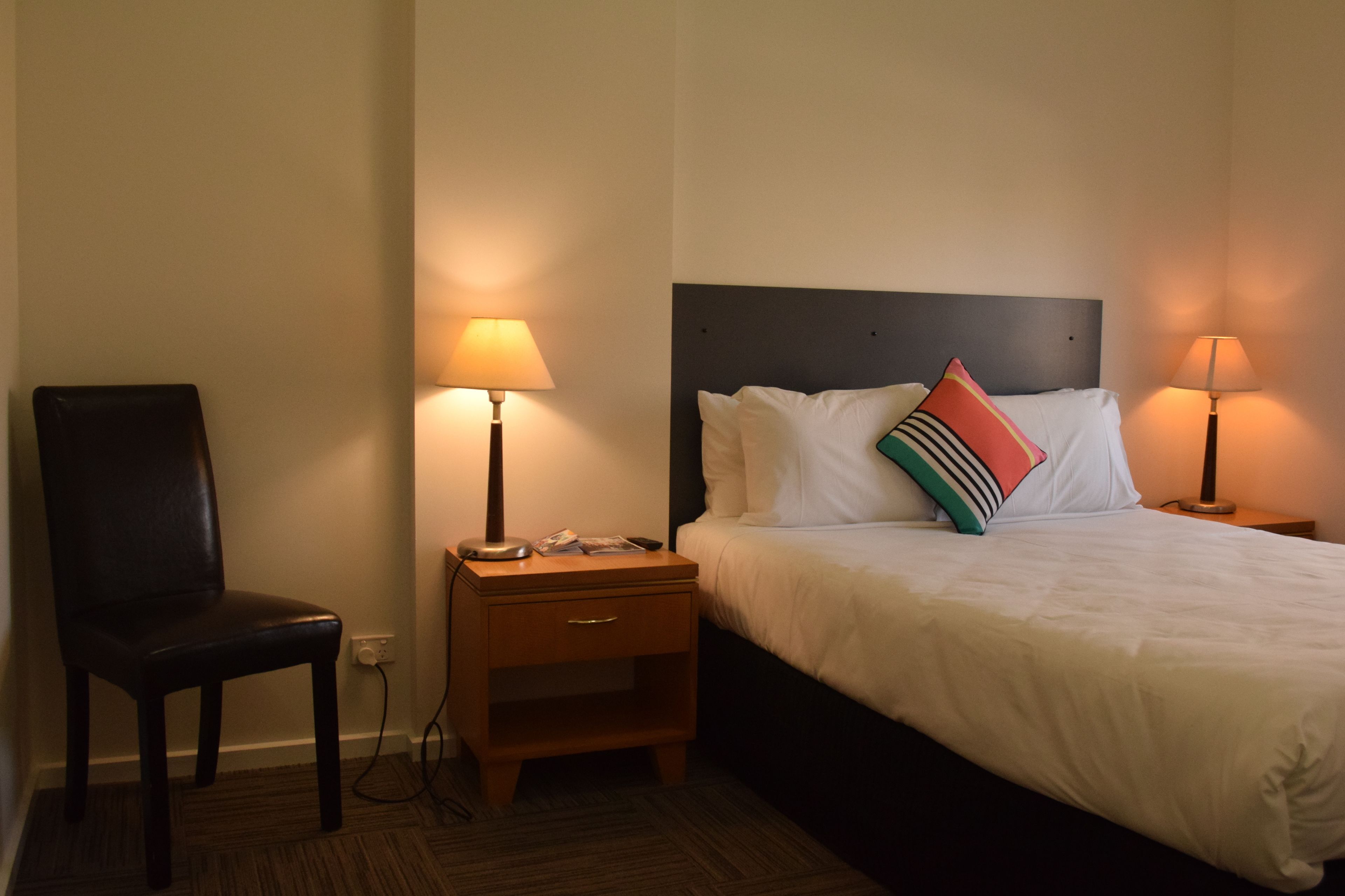 Quarto de hotel com cama, duas luminárias de cabeceira, mesa de apoio, uma cadeira e uma tomada na parede.