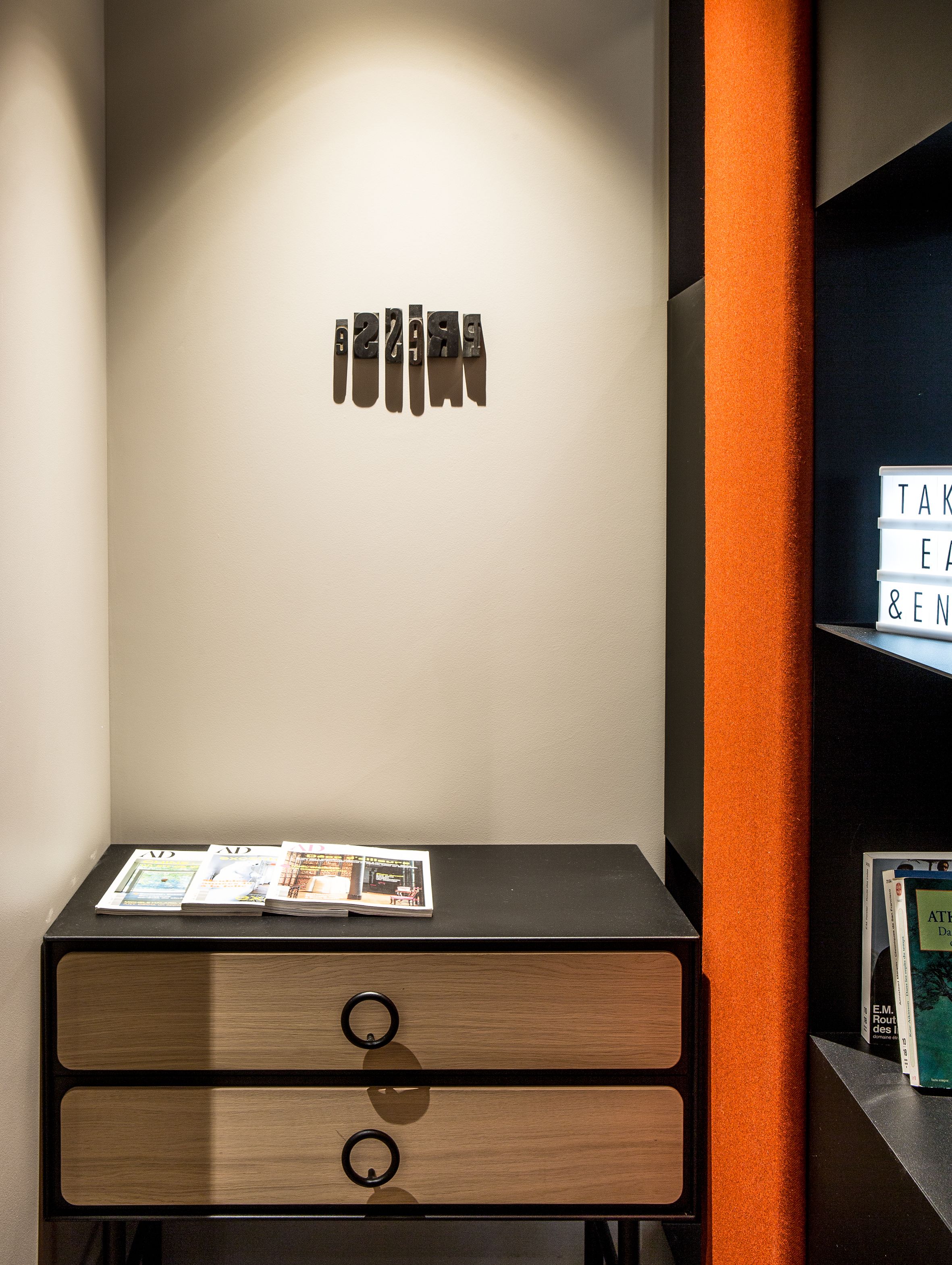 Quarto com mesa de cabeceira de madeira, uma revista e uma estante com livros e uma lightbox exibindo a frase 'TAKE ENJOY & ENJOY'.