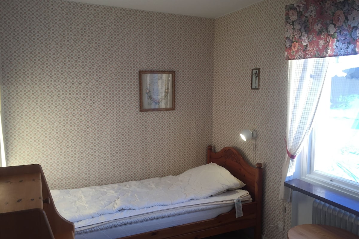 Quarto pequeno com cama de solteiro, papel de parede estampado, um certificado emoldurado na parede e uma janela com cortinas.