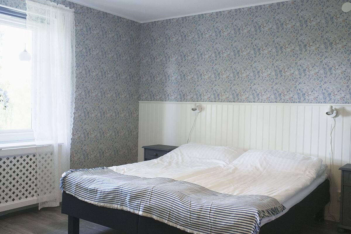 Quarto com cama, mesa de cabeceira, janela com cortinas e papel de parede floral.