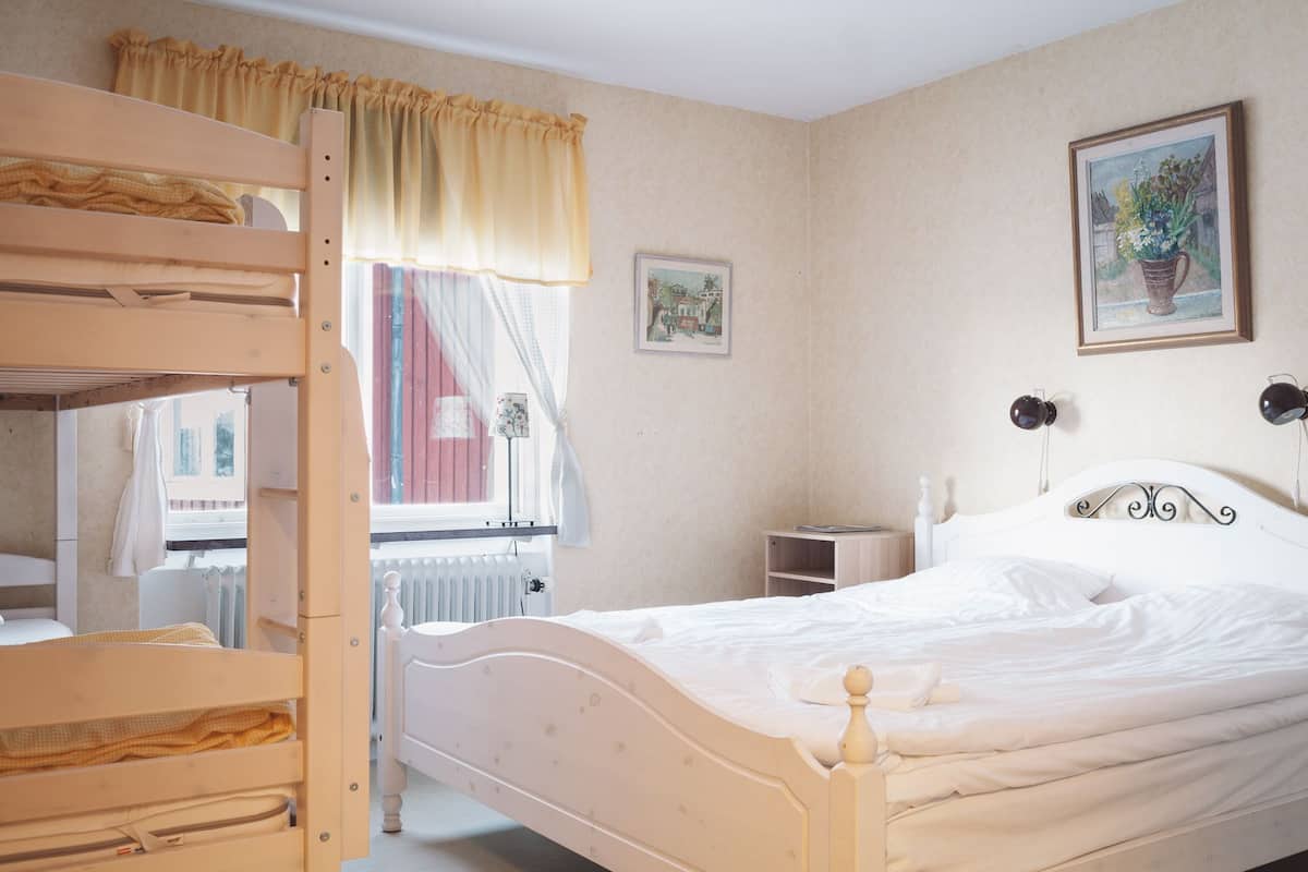 Quarto com beliche, uma cama de solteiro, radiador, janela com cortinas, mesa de cabeceira, abajur e um quadro na parede.
