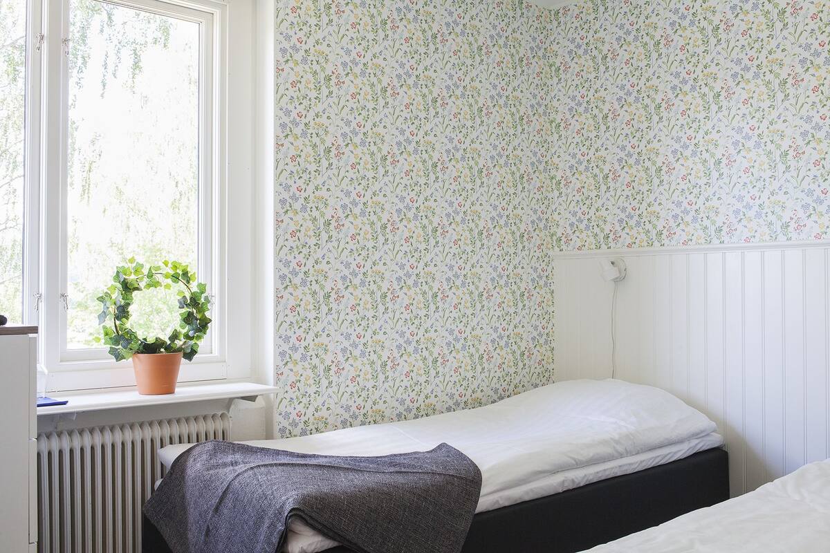 Quarto pequeno com uma cama de solteiro, radiador, janela com planta em vaso e papel de parede floral.