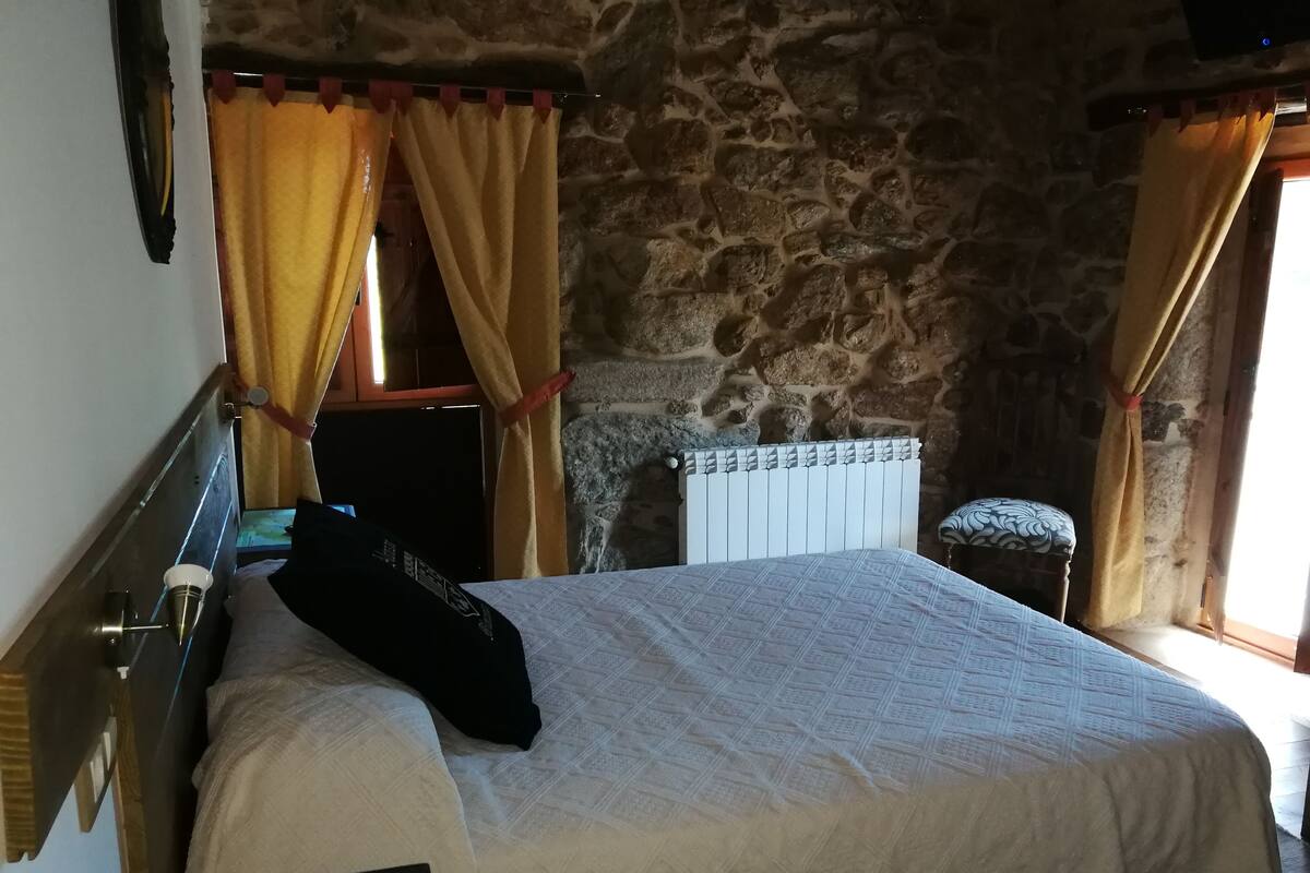 Um quarto com parede de pedra, uma cama com colcha branca, travesseiro preto e mesa de cabeceira com abajur.