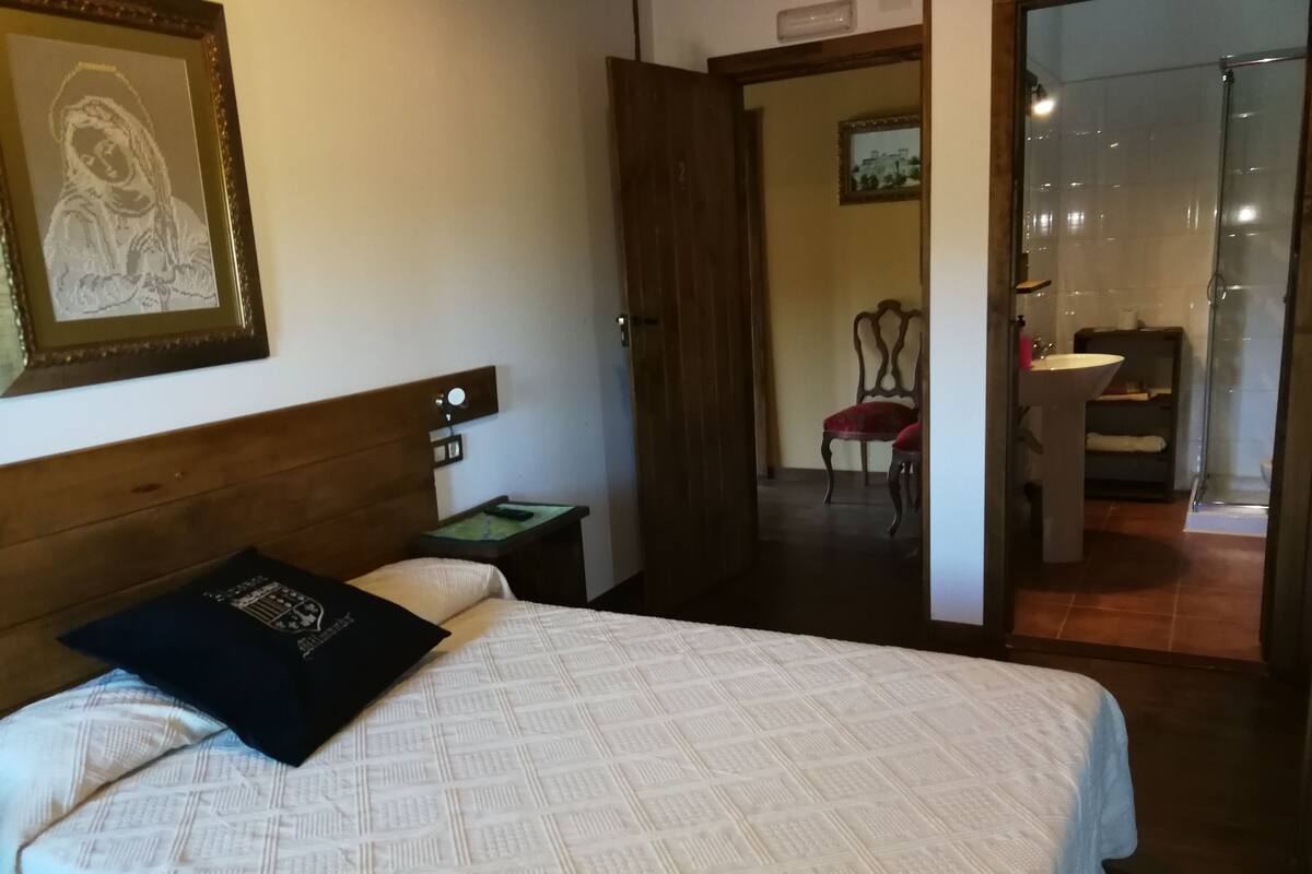 Quarto de hotel com cama, mesa de cabeceira, quadro pendurado na parede, porta de madeira e banheiro visível por uma porta aberta.