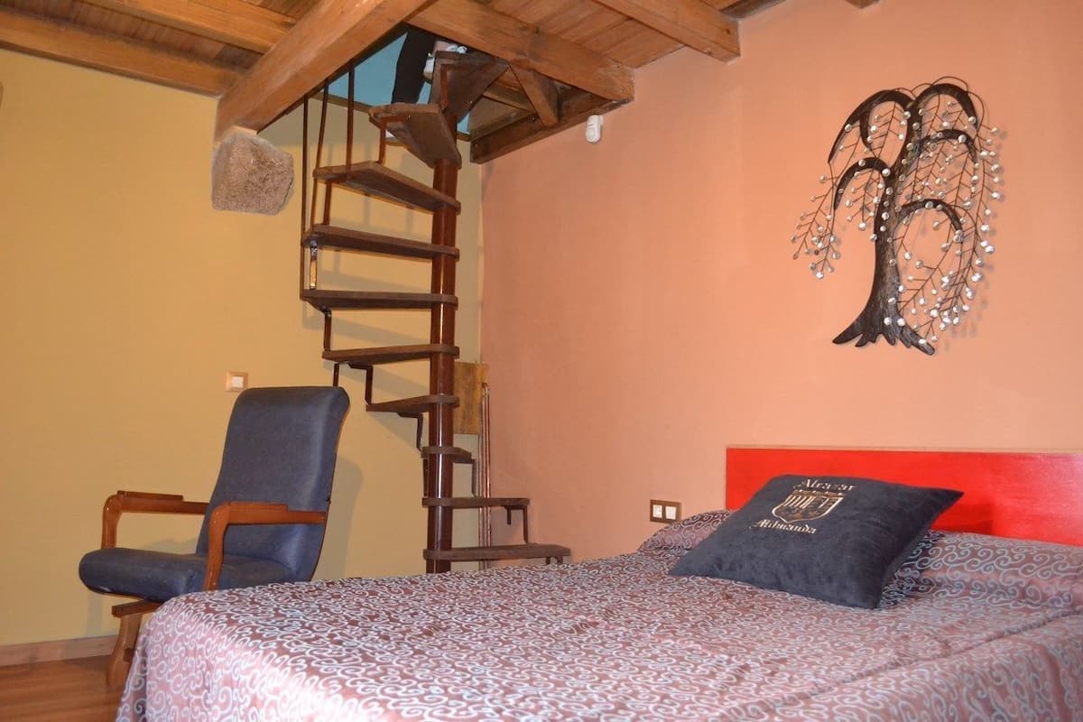Quarto com escada espiral de madeira, cama com roupa de cama estampada, uma cadeira e um objeto decorativo na parede.