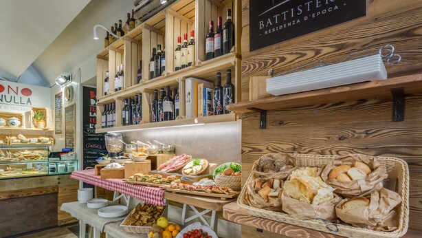 Um balcão de padaria com diversos pães e doces, uma área de exposição de vinhos com garrafas, e uma placa com a inscrição