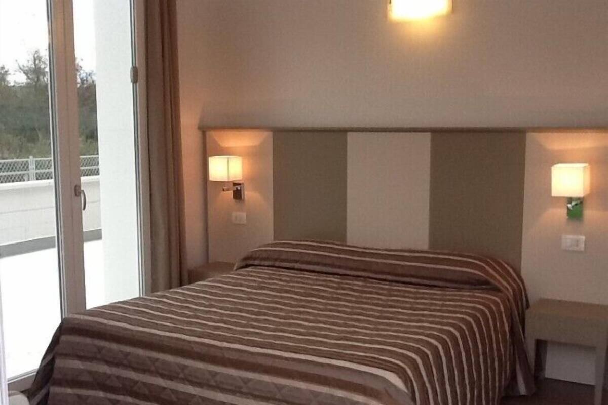 Quarto de hotel com uma cama, duas luminárias de cabeceira, uma mesinha e uma janela com cortinas.