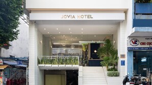 Exterior do Hotel Jovia, com fachada moderna, entrada visível e área de estacionamento com scooters.