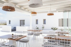 Uma área de refeições moderna ao ar livre, com móveis brancos, luminárias pendentes de vime e plantas em vasos.