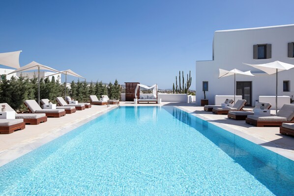 Uma área de piscina com espreguiçadeiras e guarda-sóis, cercada por edifícios brancos e cactos.