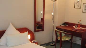 Quarto de hotel com cama, escrivaninha, cadeira, luminária de mesa, telefone e espelho.