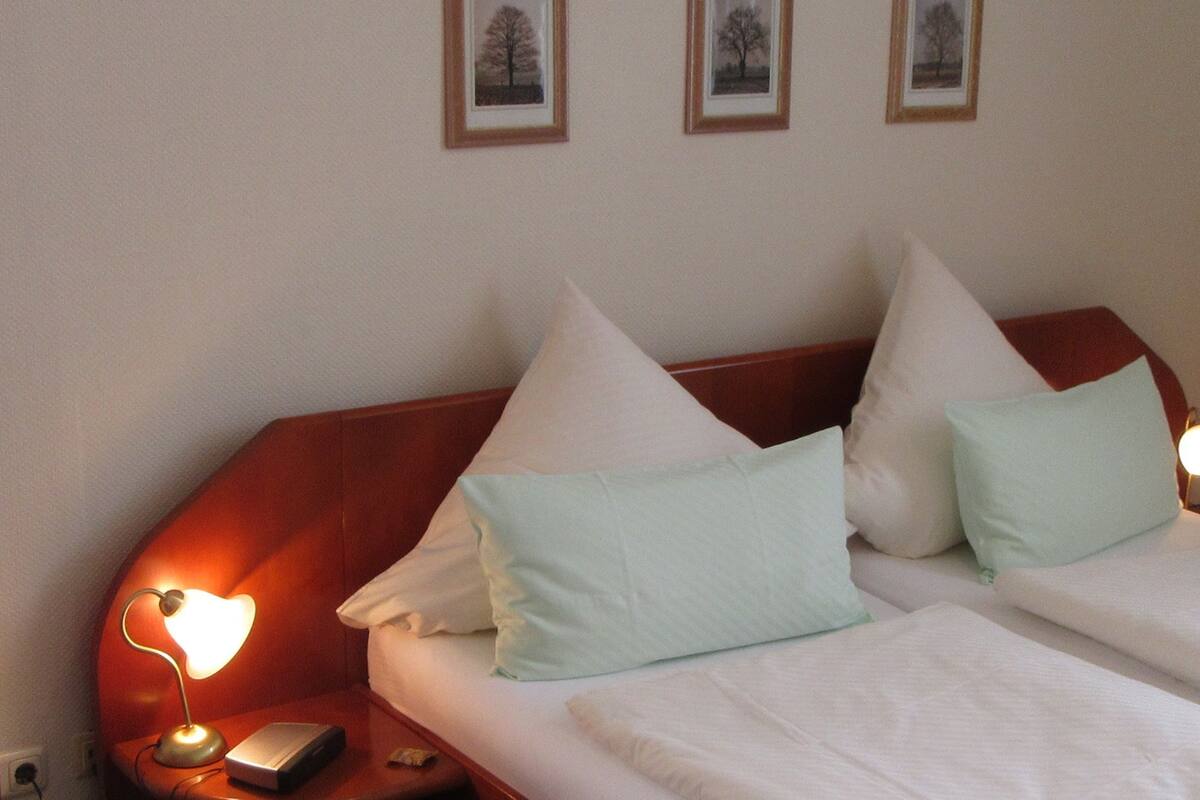 Quarto de hotel com cabeceira de madeira, duas camas com roupa de cama branca e almofadas verde-claras, abajur, pequena mesa com luminária e controle remoto, e três quadros na parede.