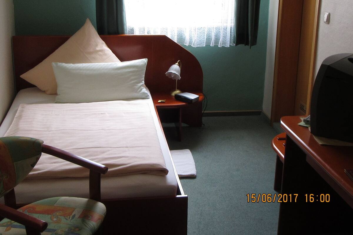 Quarto de hotel com cama, uma cadeira, escrivaninha com luminária e televisão.