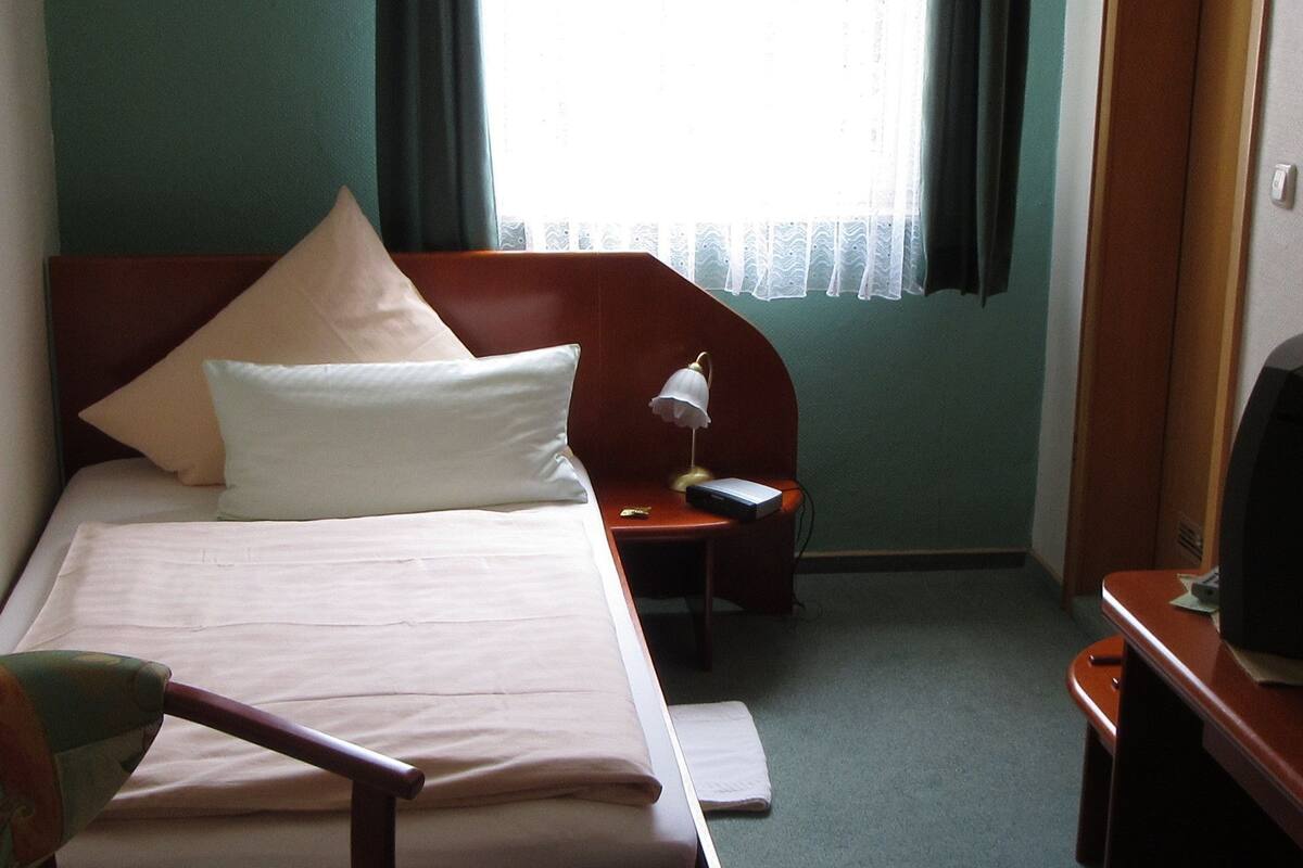 Quarto de hotel com cama, mesa de cabeceira com abajur, cadeira, televisão e janela com cortinas.