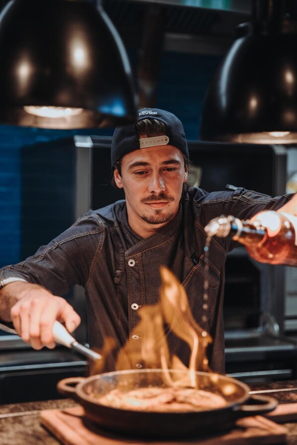 Um cozinheiro preparando comida em uma panela sobre o fogão, com uma garrafa de molho, em um ambiente de cozinha.