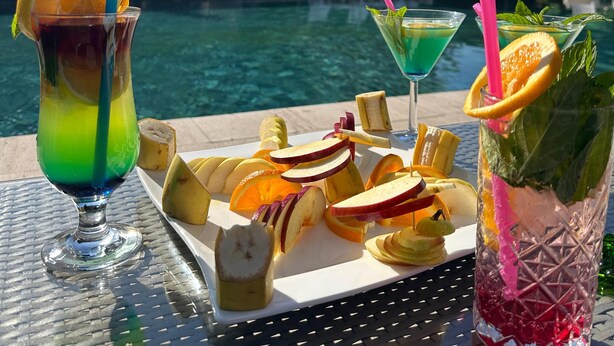 Mesa à beira da piscina com três bebidas e um prato de frutas.
