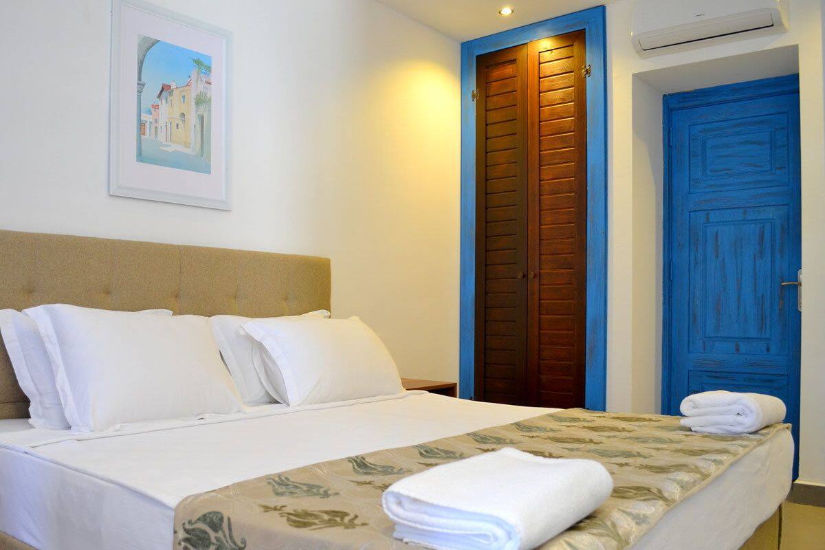 Uma cama bem arrumada com lençóis brancos e um edredom estampado com flores, uma cabeceira de madeira e uma porta azul ao fundo.