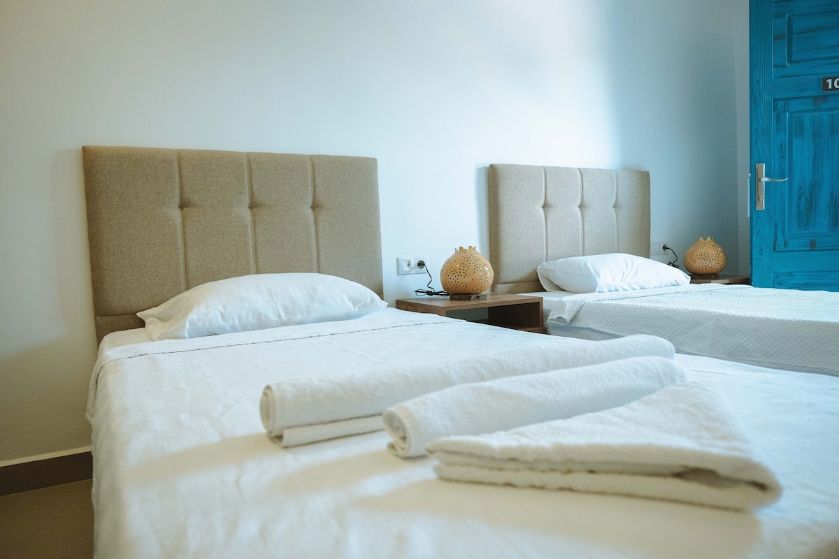 Quarto de hotel com duas camas, cada uma com cabeceira e roupa de cama branca. Há duas mesas de cabeceira com abajures e objetos decorativos.
