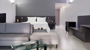 Quarto de hotel moderno com um sofá cinzento, uma mesa de centro de vidro, uma cama com roupa de cama branca, uma mesa de cabeceira com abajur e uma televisão sobre um suporte.