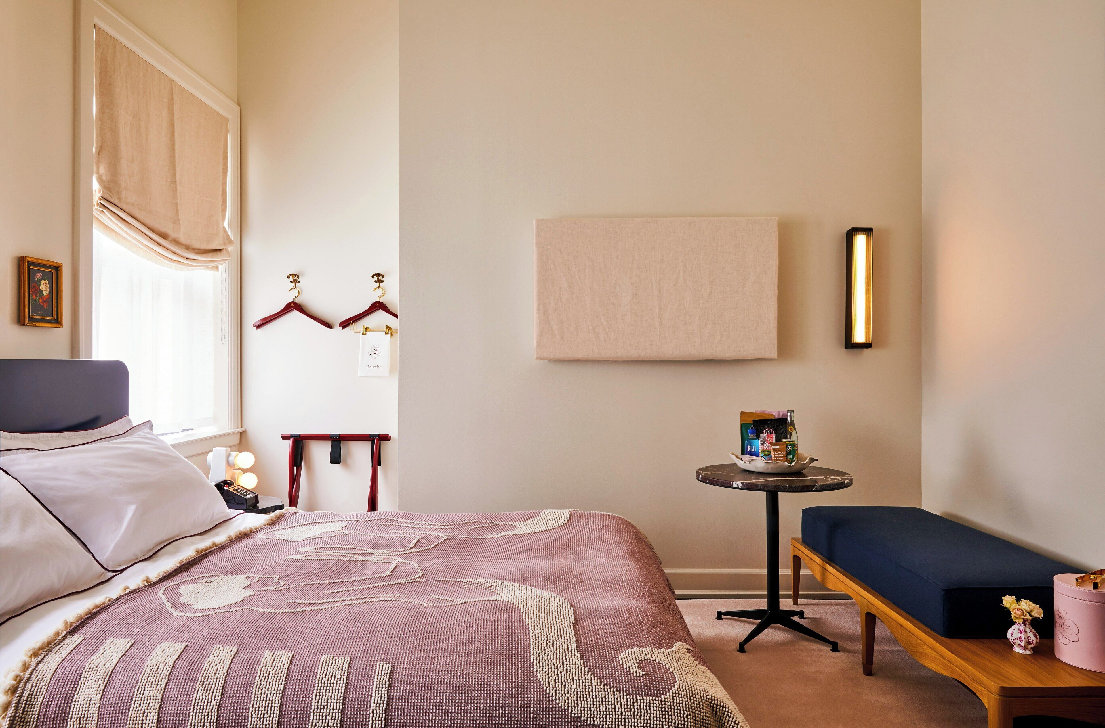 Quarto bem arrumado com cama, mesa de cabeceira, banqueta e uma pequena mesa com um vaso.
