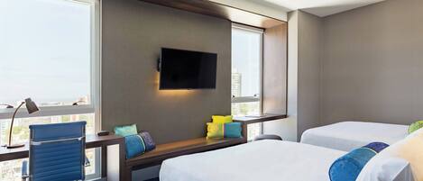 Quarto de hotel com uma janela grande, uma escrivaninha com televisão de tela plana, duas camas e uma cadeira.