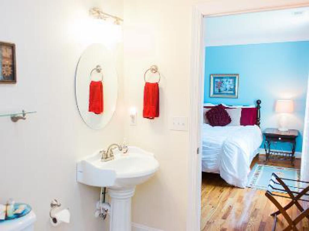 Casa de banho com lavatório, espelho e sanita. O quarto tem uma cama com roupa de cama branca e almofada vermelha. Há uma mesa de cabeceira com uma lâmpada e um porta-retrato na parede.